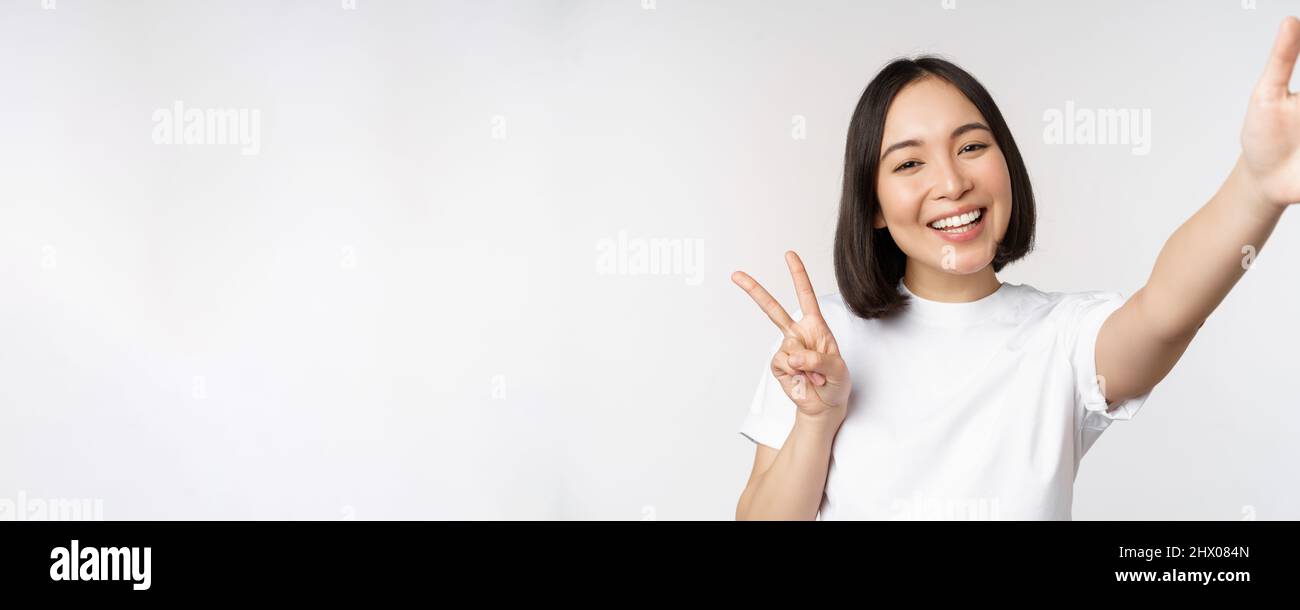 Beautiful young asian woman taking selfie, posing with peace v-sign, smiling happy, take photo ...