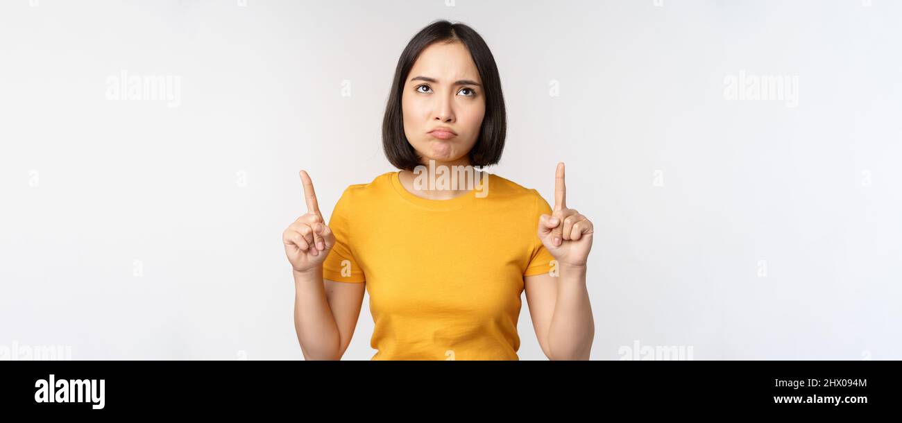 Disappointed asian woman looking, pointing fingers up with angry moody ...