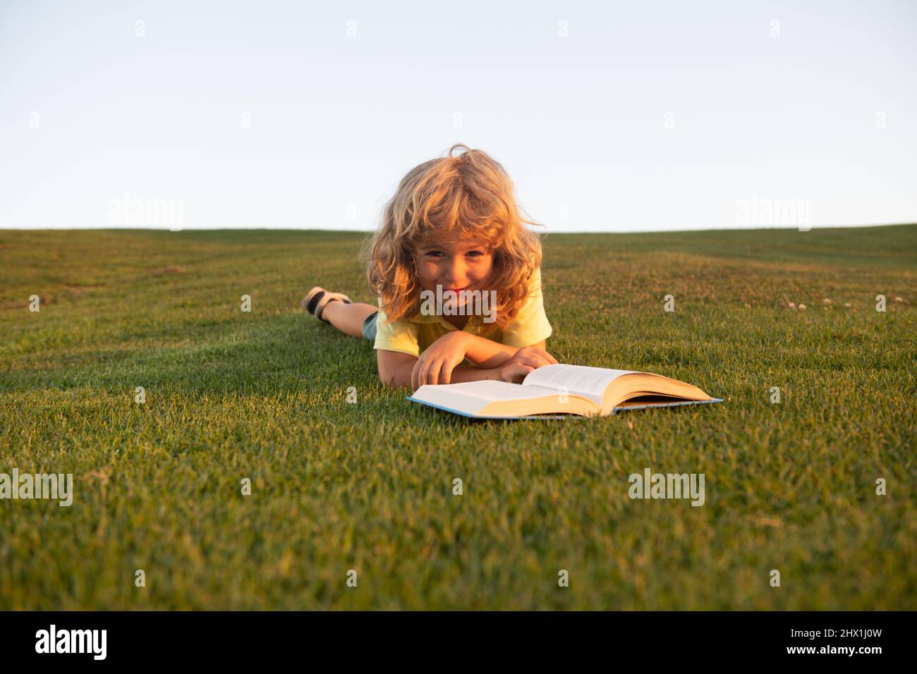 Clever child boy reading book laying on grass on grass and sky ...