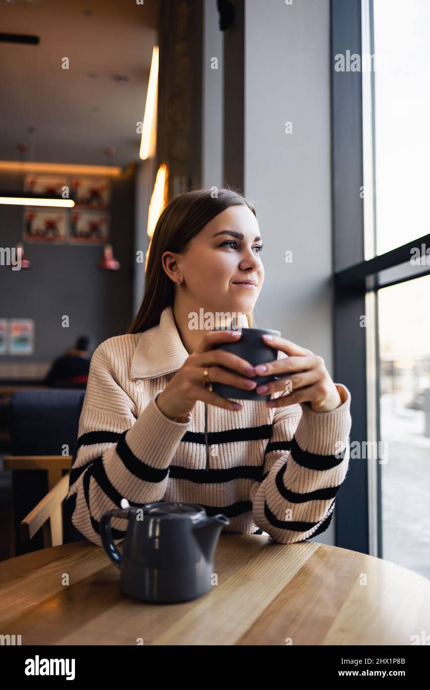 A young beautiful woman drinks coffee and looks out the window while ...