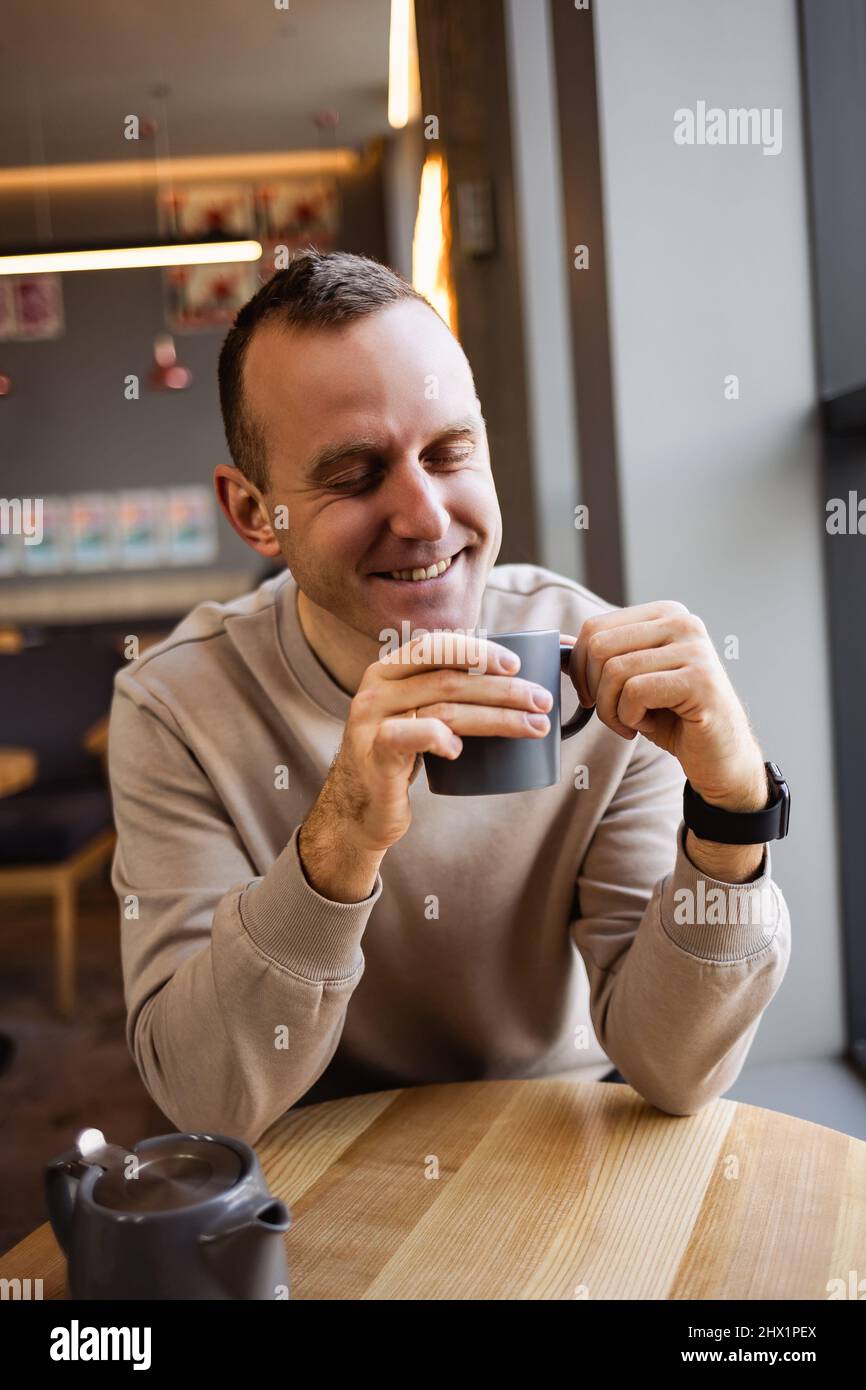 A handsome smiling calm young man sits in a cafe at a table drinking ...