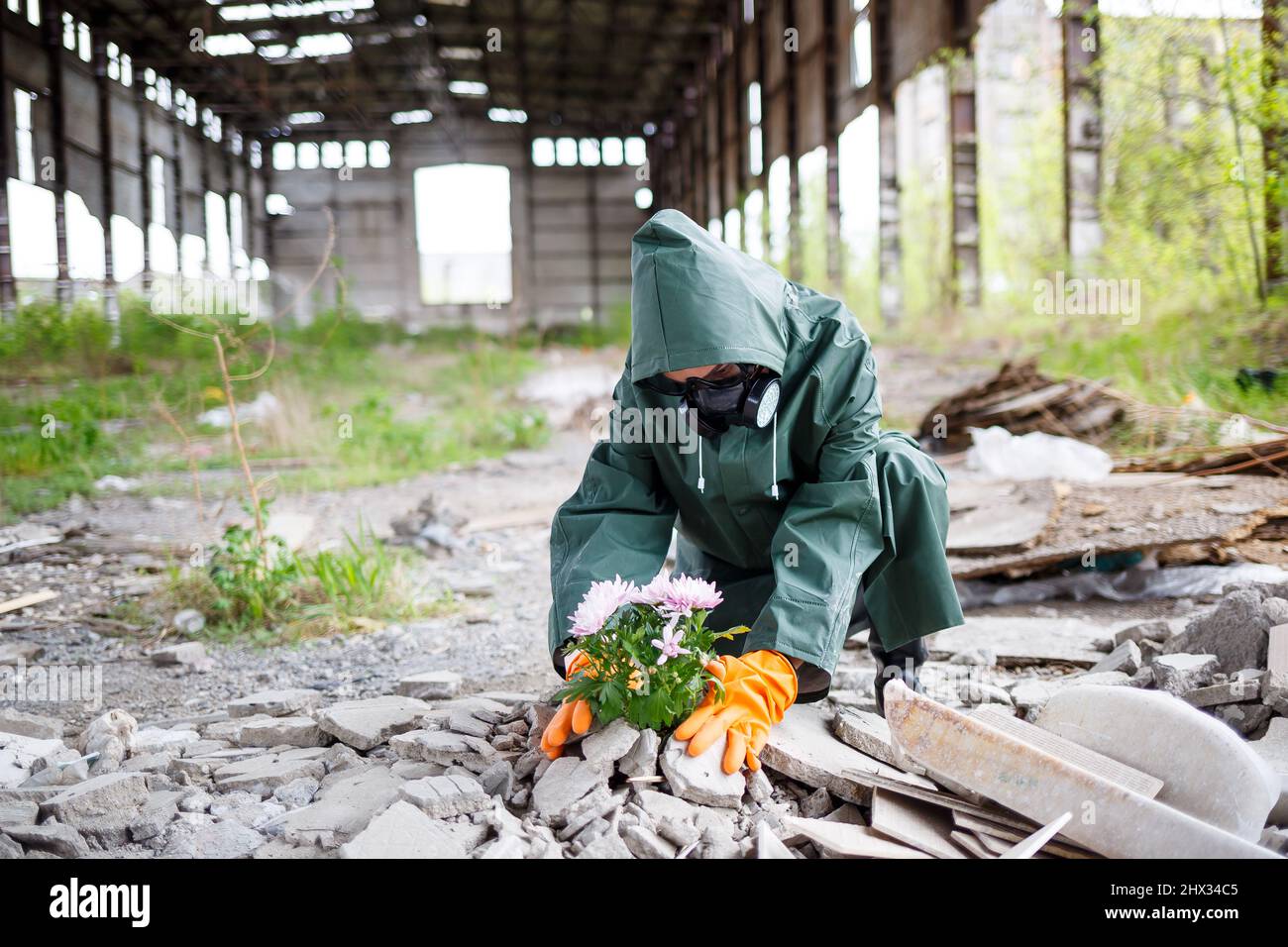 A man in a raincoat and gas mask collects a flower from a scorched ...