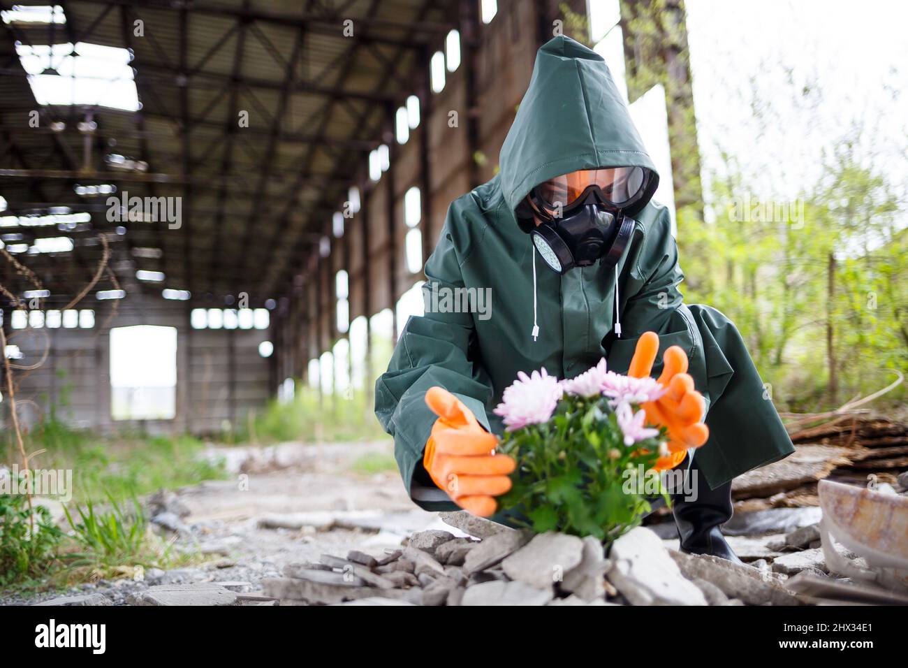 A man in a raincoat and gas mask collects a flower from a scorched ...