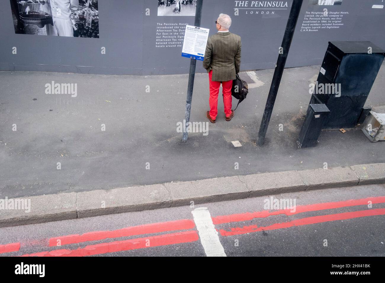 A man wearing red trousers pauses to read a construction hoarding, near ...