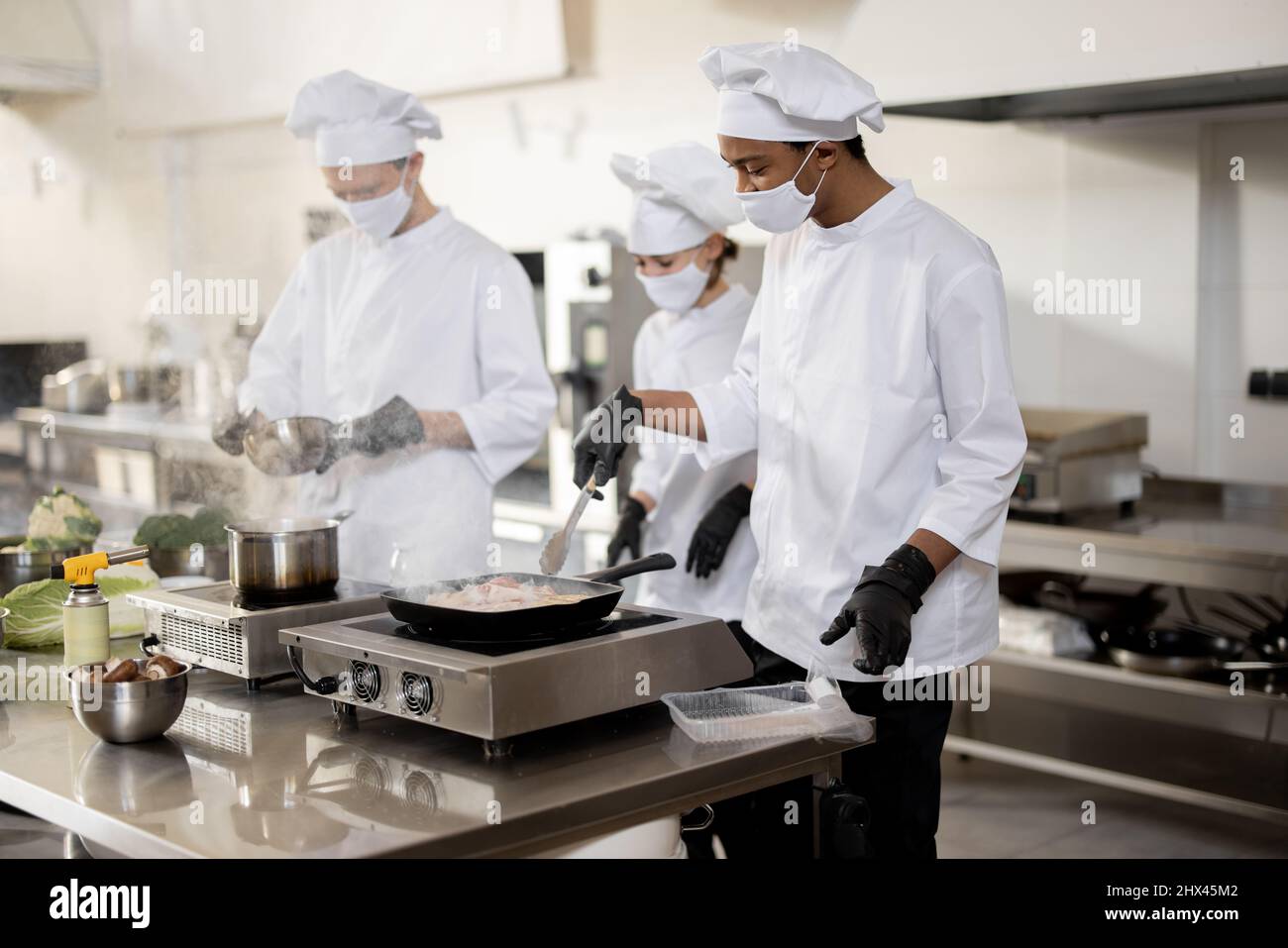 Multiracial team of cooks in uniform and face masks cooking meals for a ...