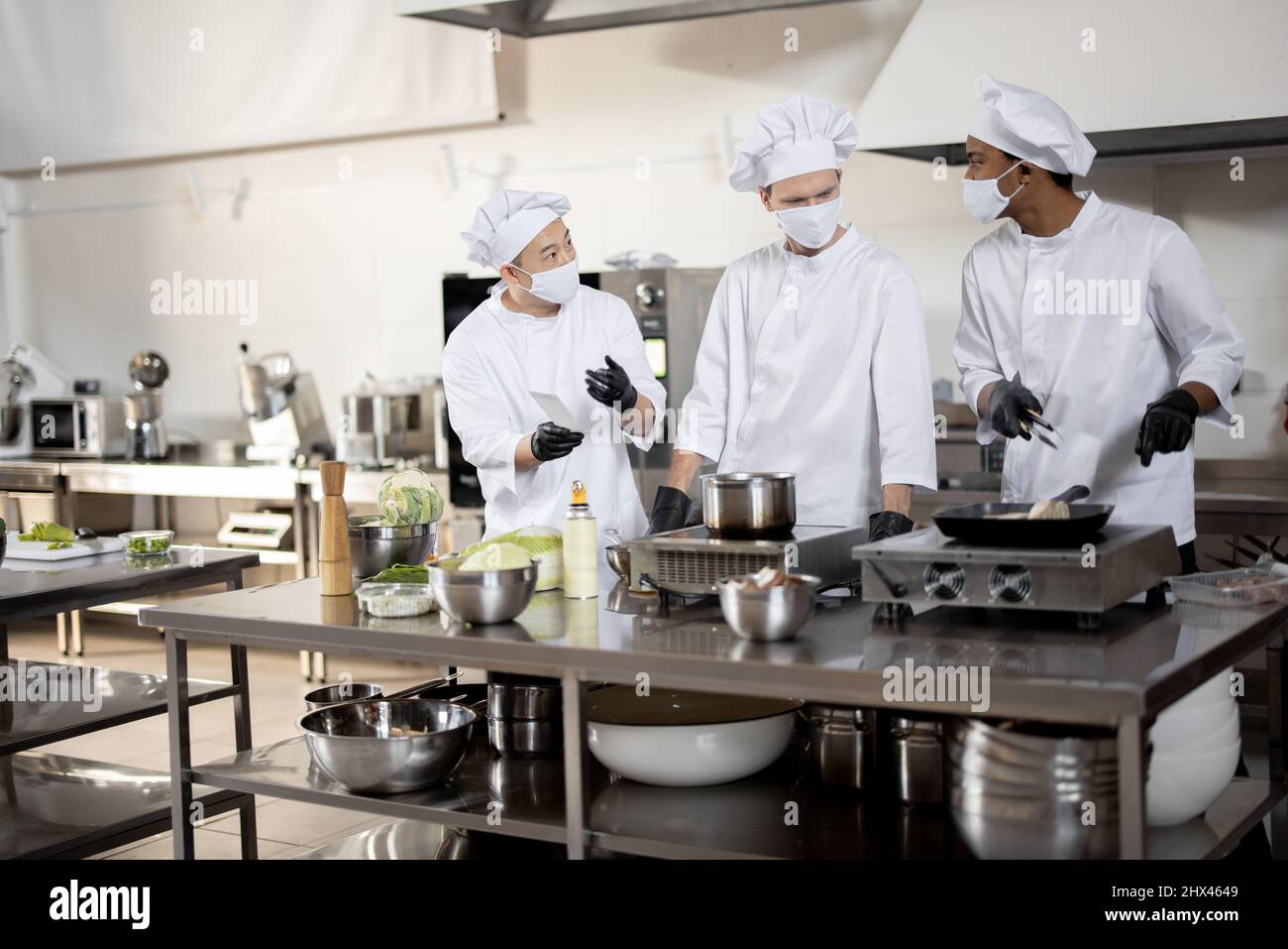 Multiracial team of cooks in uniform and face masks cooking meals for a ...