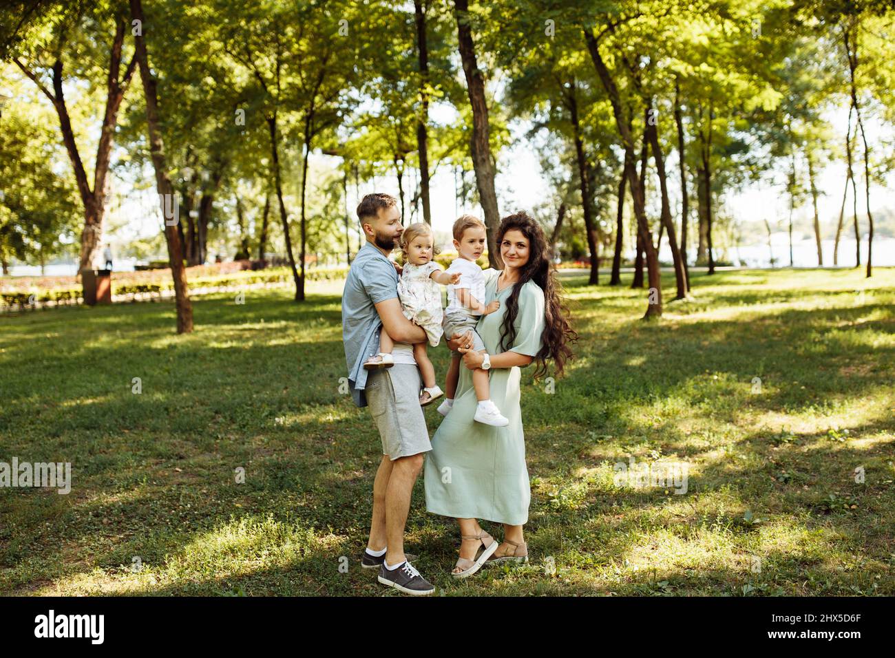Overjoyed parents with little children walking at the park, lovely mom hold in arms joyful son ...