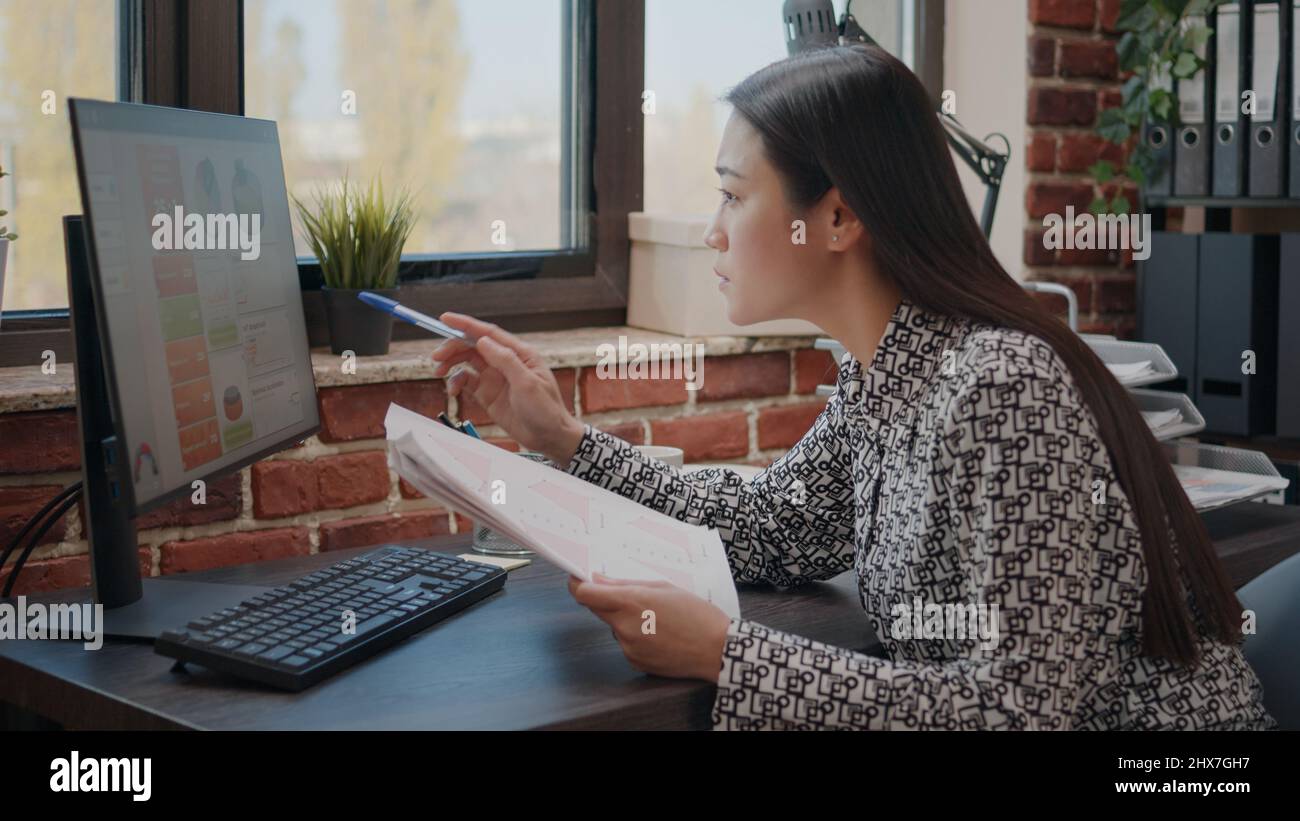 Asian woman comparing charts on papers and computer, working to plan business project in startup ...