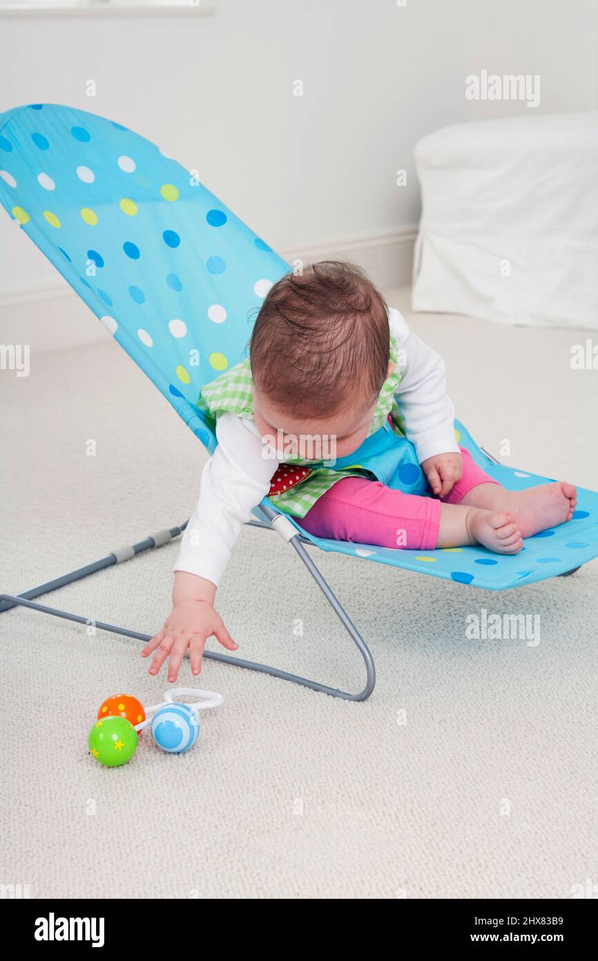 Baby girl sat in baby chair reaching for colourful plastic rattle toy ...