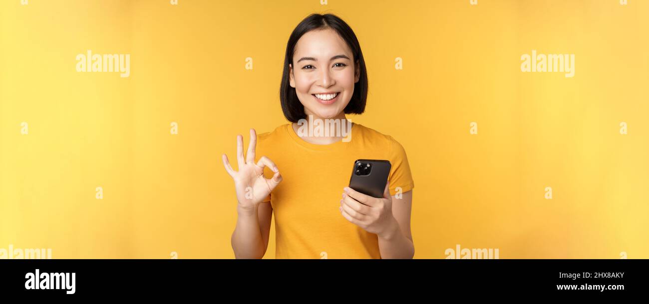 Happy smiling asian girl holding mobile phone and showing okay ...