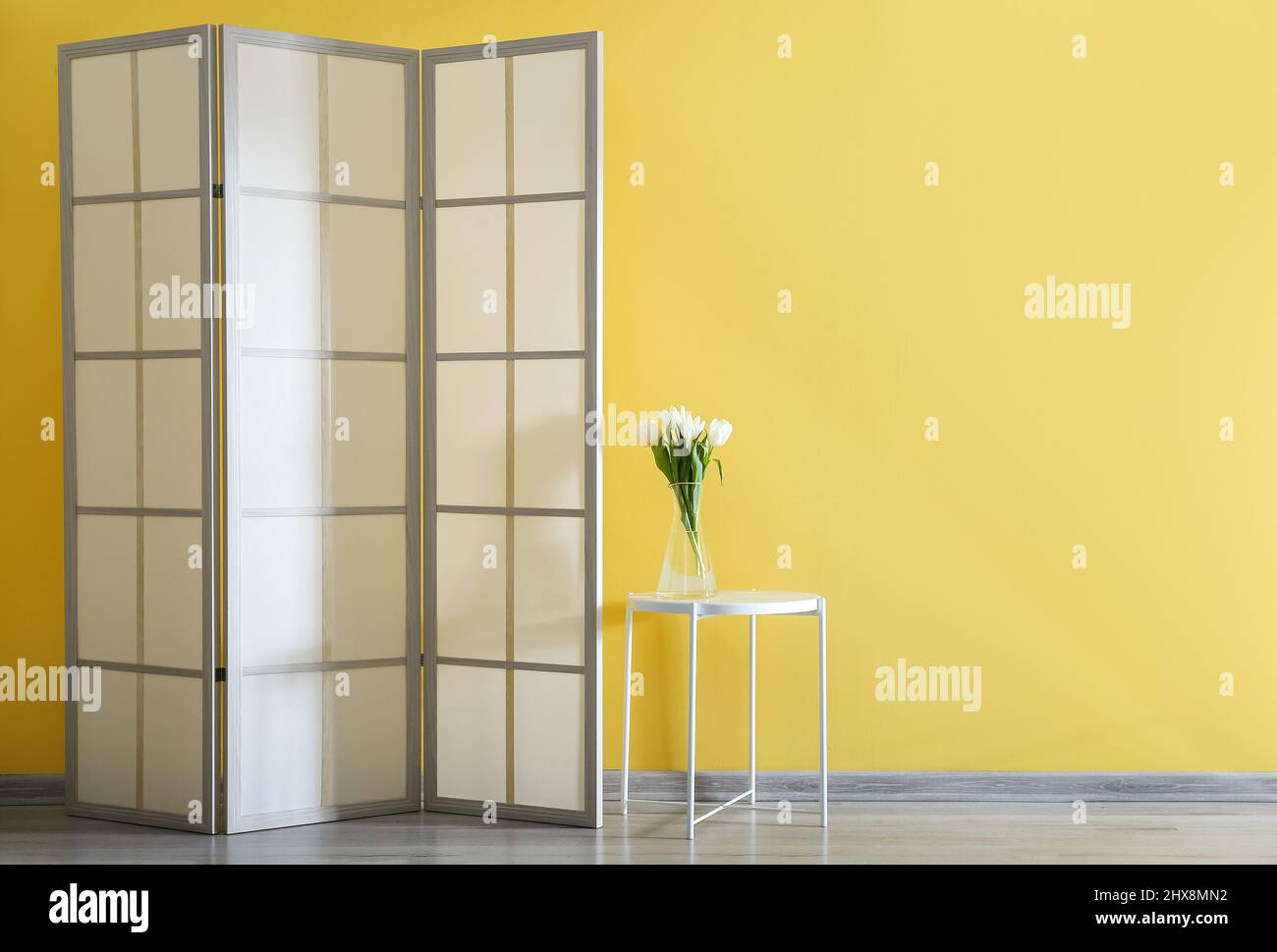 Stylish folding screen and vase with flowers on table near color wall ...