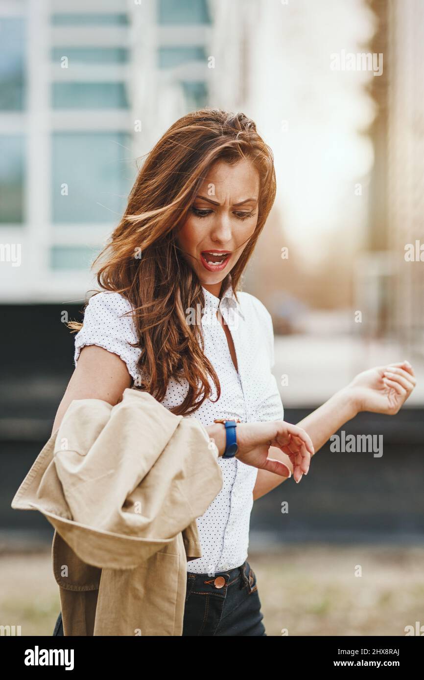 Beautiful young woman worriedly looking on the watch against urban city ...