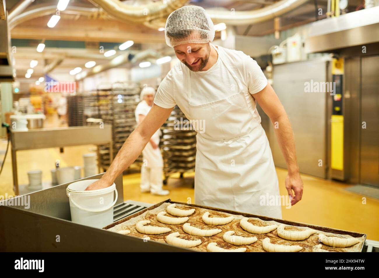 Young man apprentice baker sprinkles croissants with poppy seeds in ...