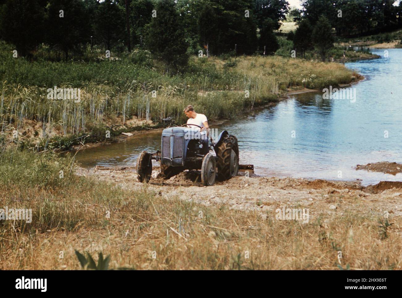 Man working on a tractor on the shore of a lake ca. 1960 Stock Photo ...