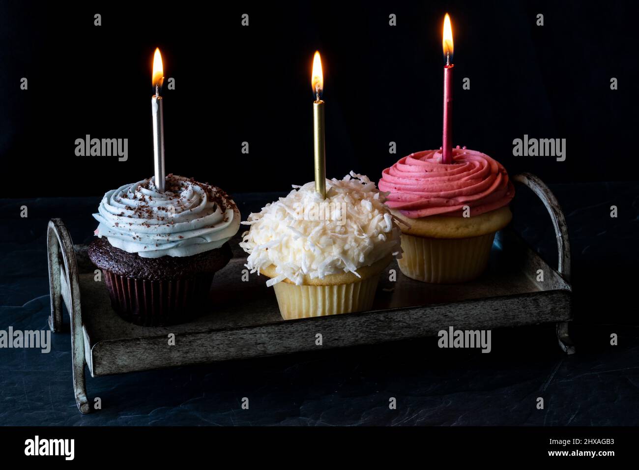 Three cupcakes with lit candles on a metal tray, against a dark ...