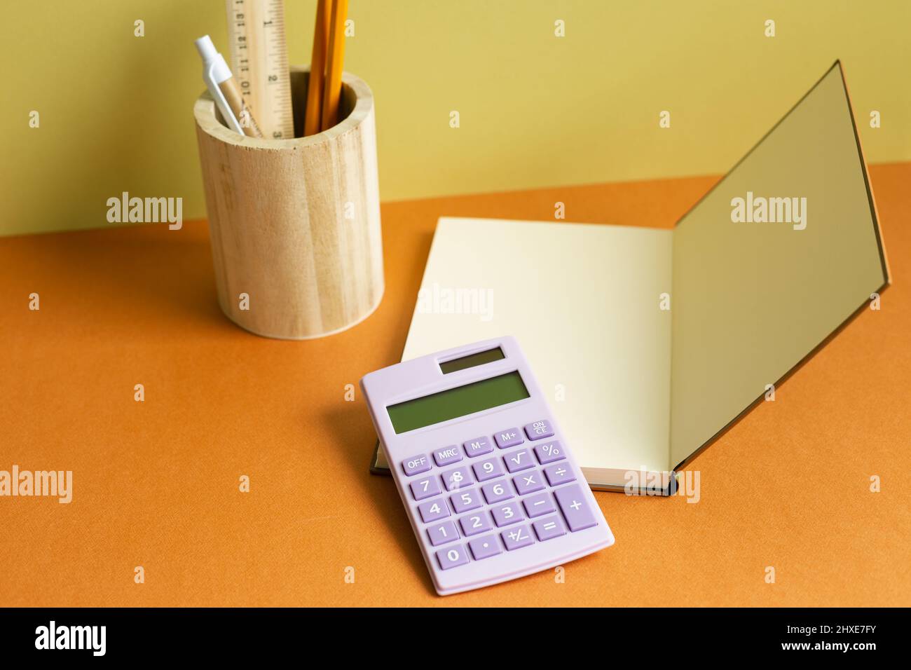 Notebook, calculator, pencil holder on orange desk. yellow wall ...