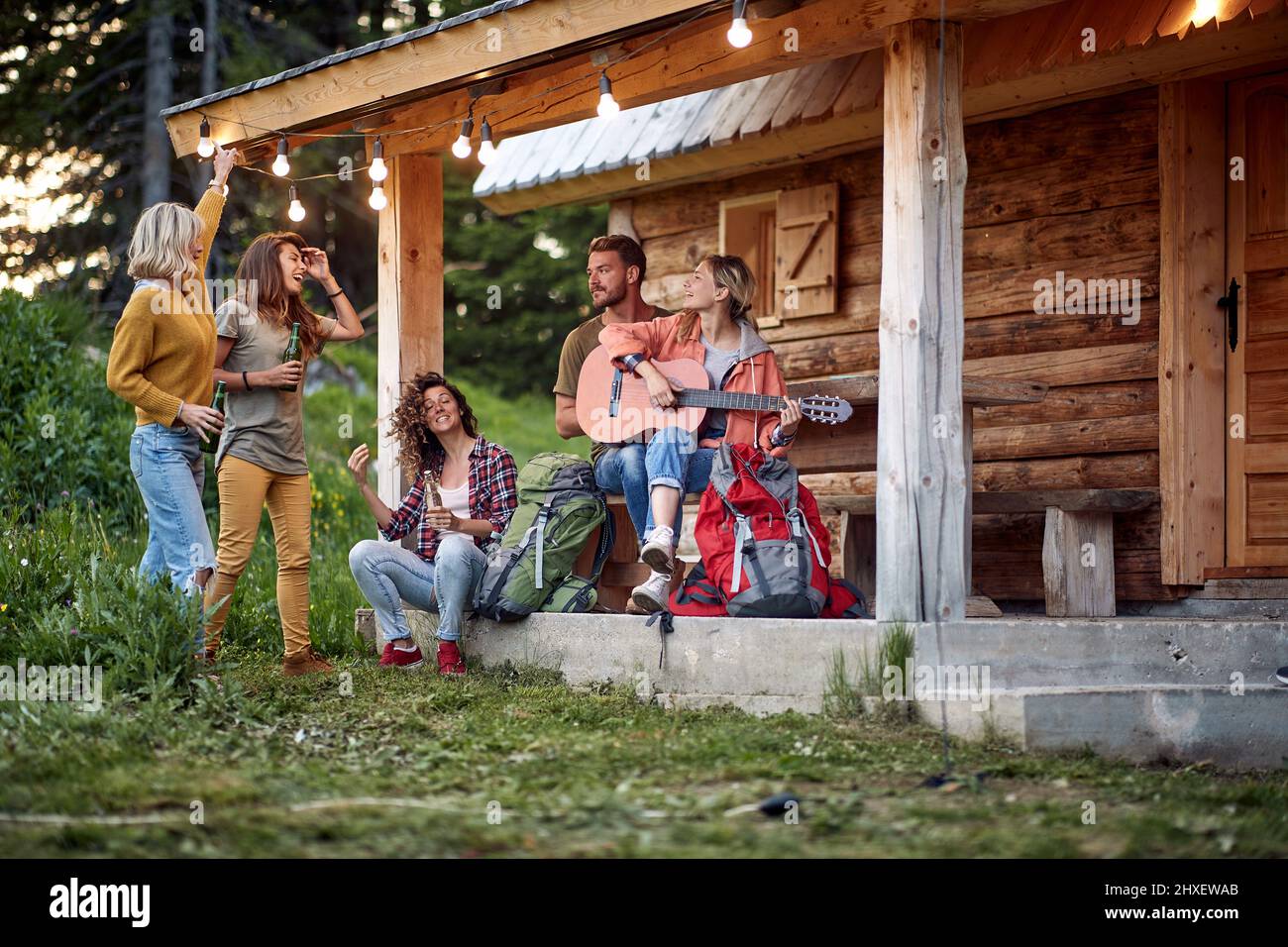 Smiling friends drinking and playing guitar in front of wooden cottage ...