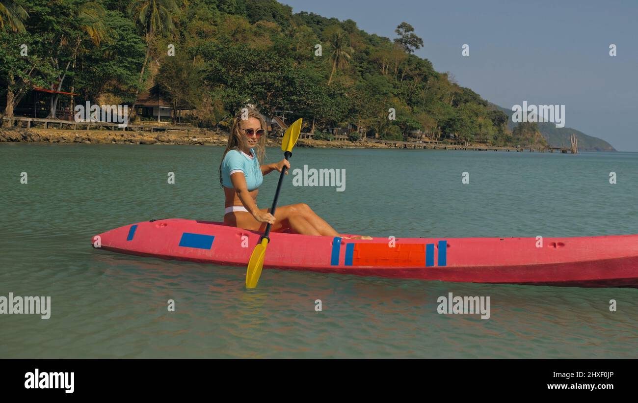 Young blonde woman in blue swimsuit rows pink plastic canoe along azure ...