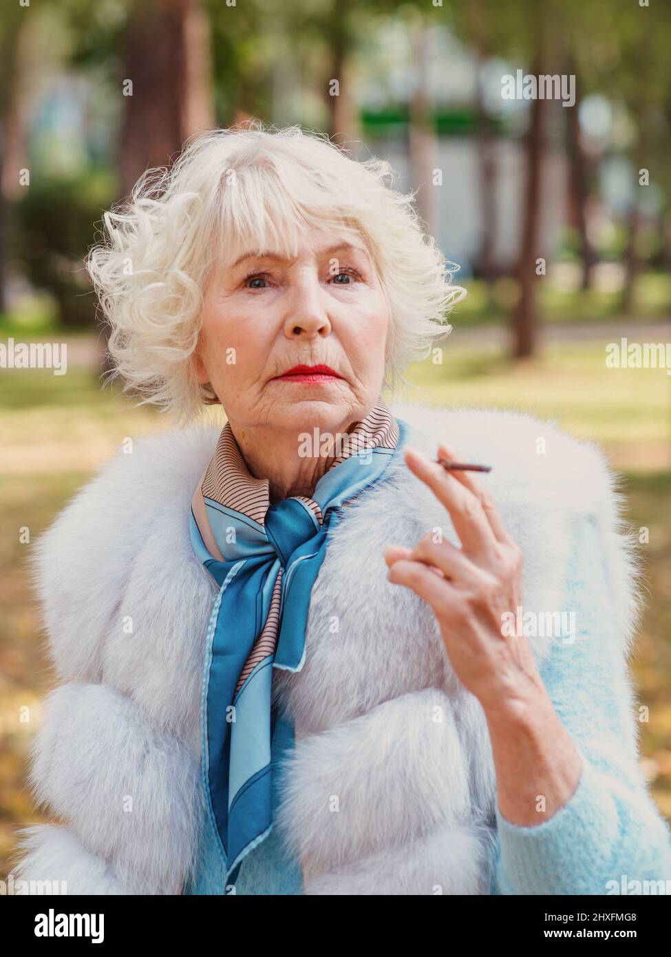 sad fashionable senior woman with grey hair in fur coat outdoor smoking ...