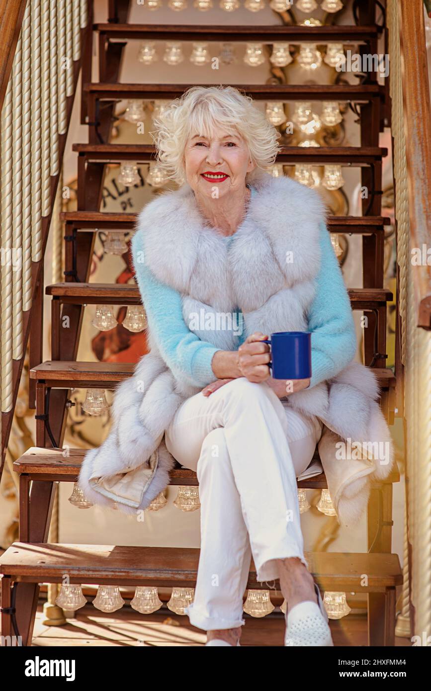 Senior Stylish Woman In Fur Coat And With Grey Hair Sitting On Carousel senior-stylish-woman-in-fur-coat-and-with-grey-hair-sitting-on-carousel