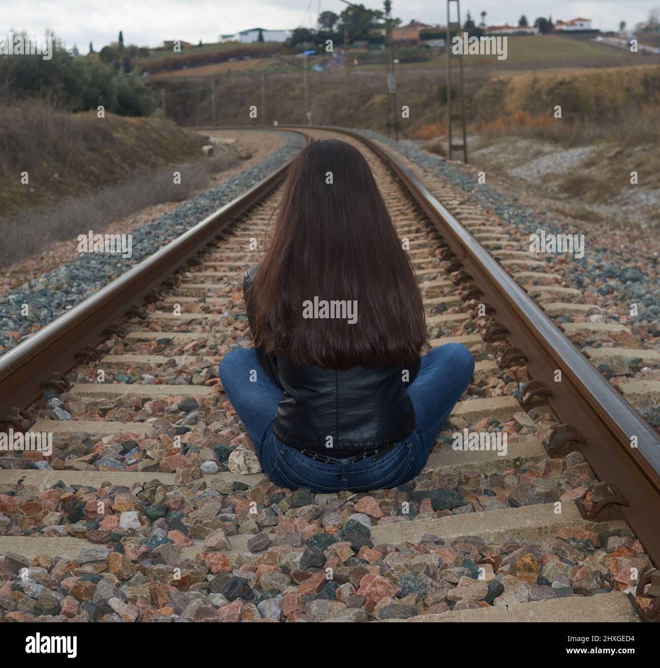 A young girl sitting on train rails and with her back to the camera ...