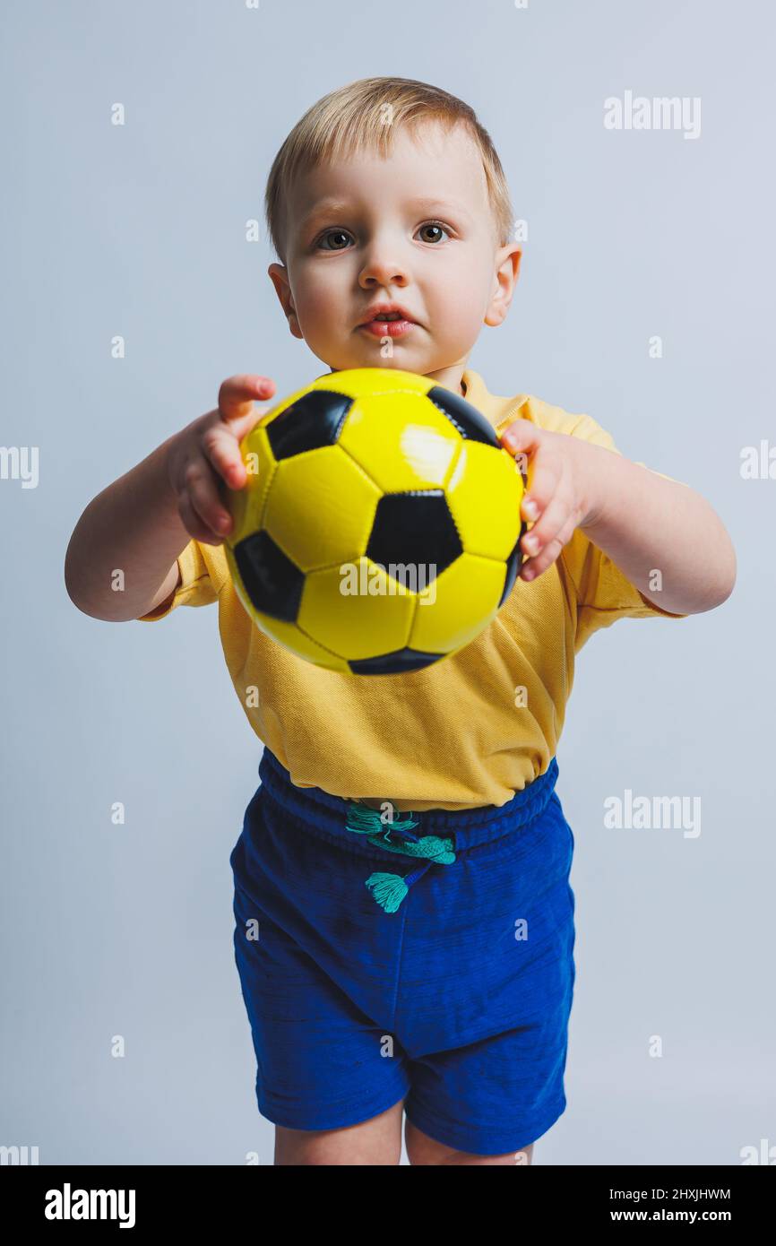 A little boy in a yellow t-shirt with a soccer ball in his hand smiles ...
