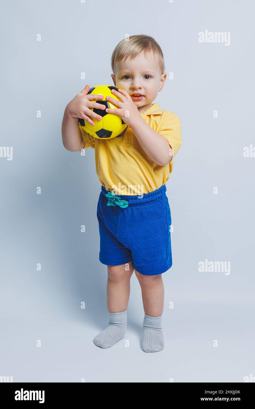 A little boy in a yellow t-shirt with a soccer ball in his hand smiles ...