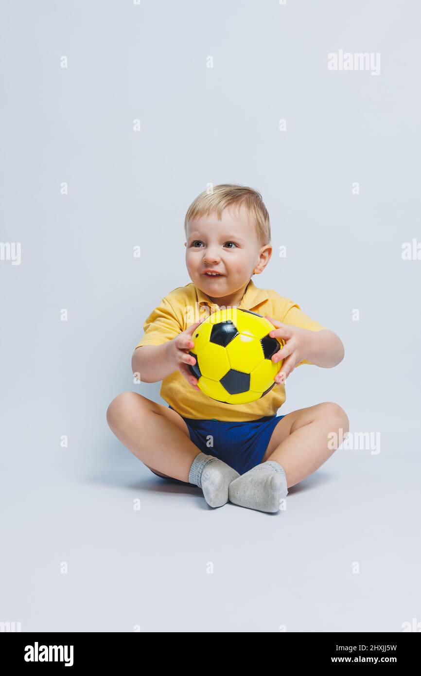 A little boy in a yellow t-shirt with a soccer ball in his hand smiles ...