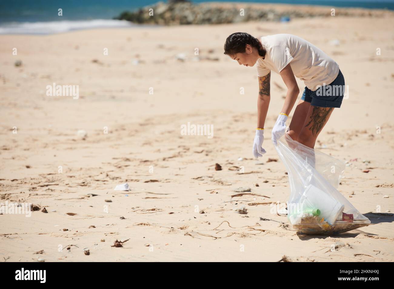 Young woman cleaning up sea beach, collecting trash into plastic bag ...