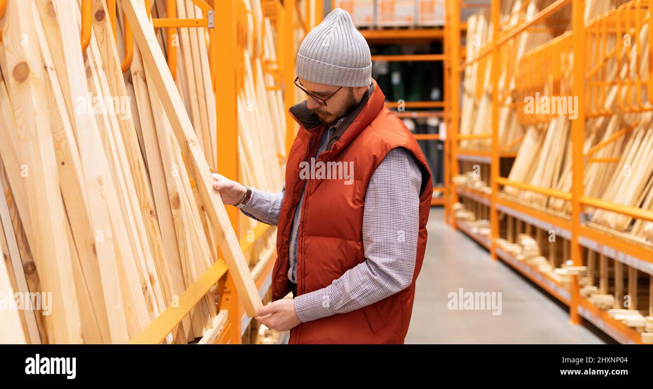 a male builder in a store chooses lumber among wooden bars and planks ...