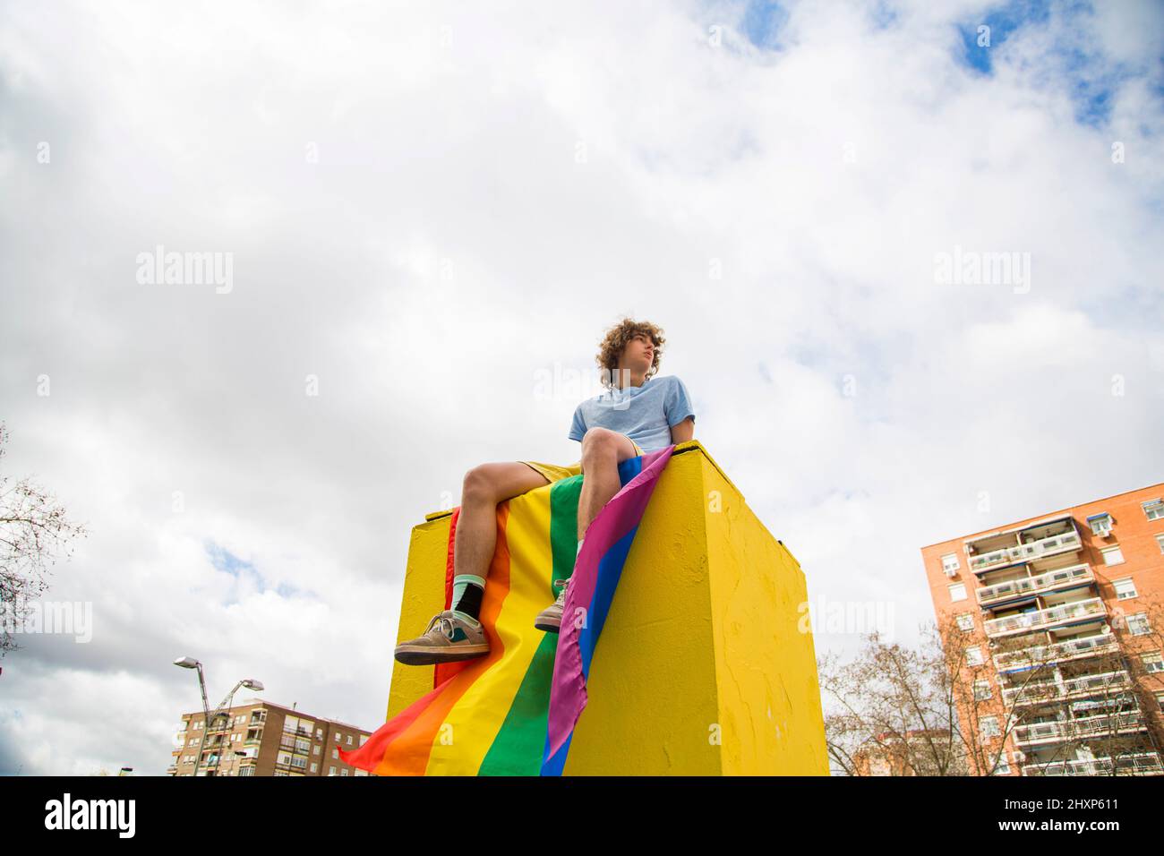 young man with curly hair sitting on a gay pride flag in town Stock ...