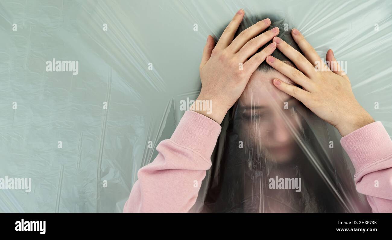 young woman with distorted face behind plastic film grabs her head ...