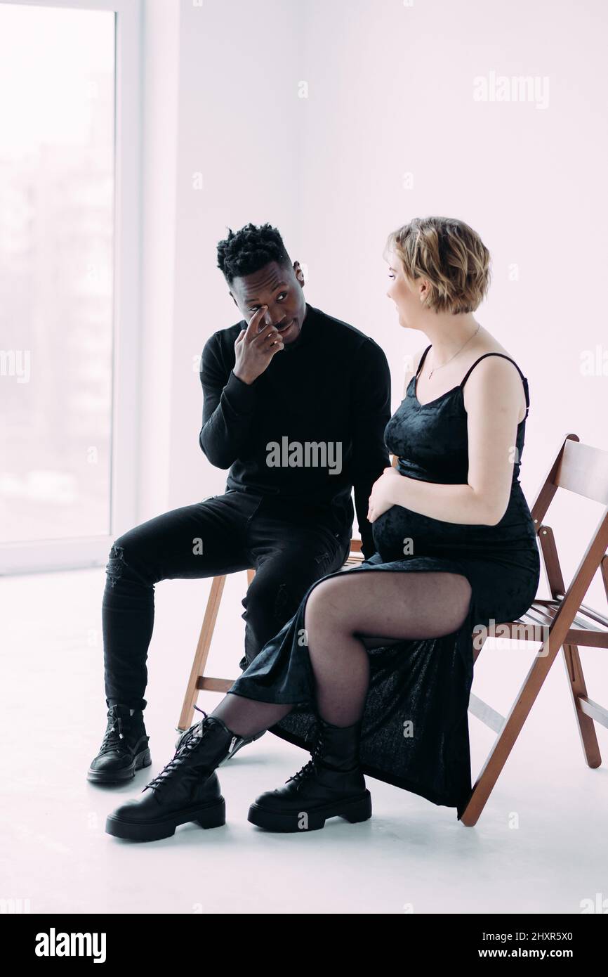 Young african man and his caucasian pregnant woman sit on chairs and ...