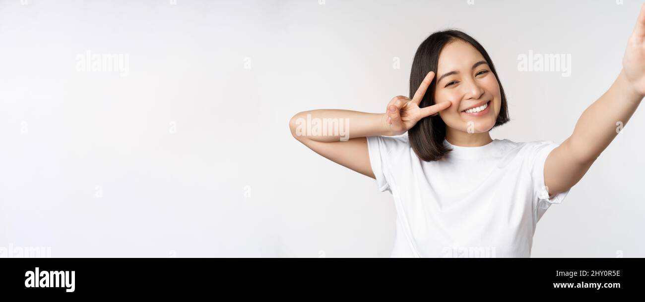 Beautiful young asian woman taking selfie, posing with peace v-sign, smiling happy, take photo ...