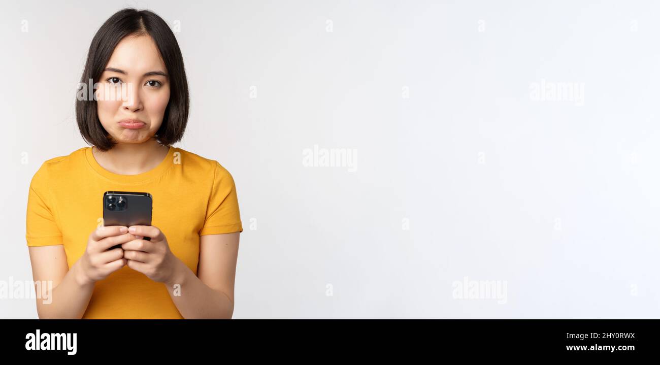 Sad asian woman holding smartphone, looking upset with regret, standing ...