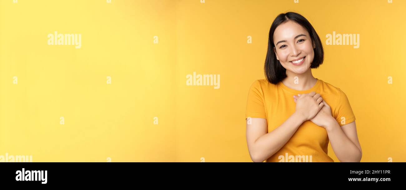 Romantic korean girl, asian woman holding hands on heart, smiling with care and tenderness ...