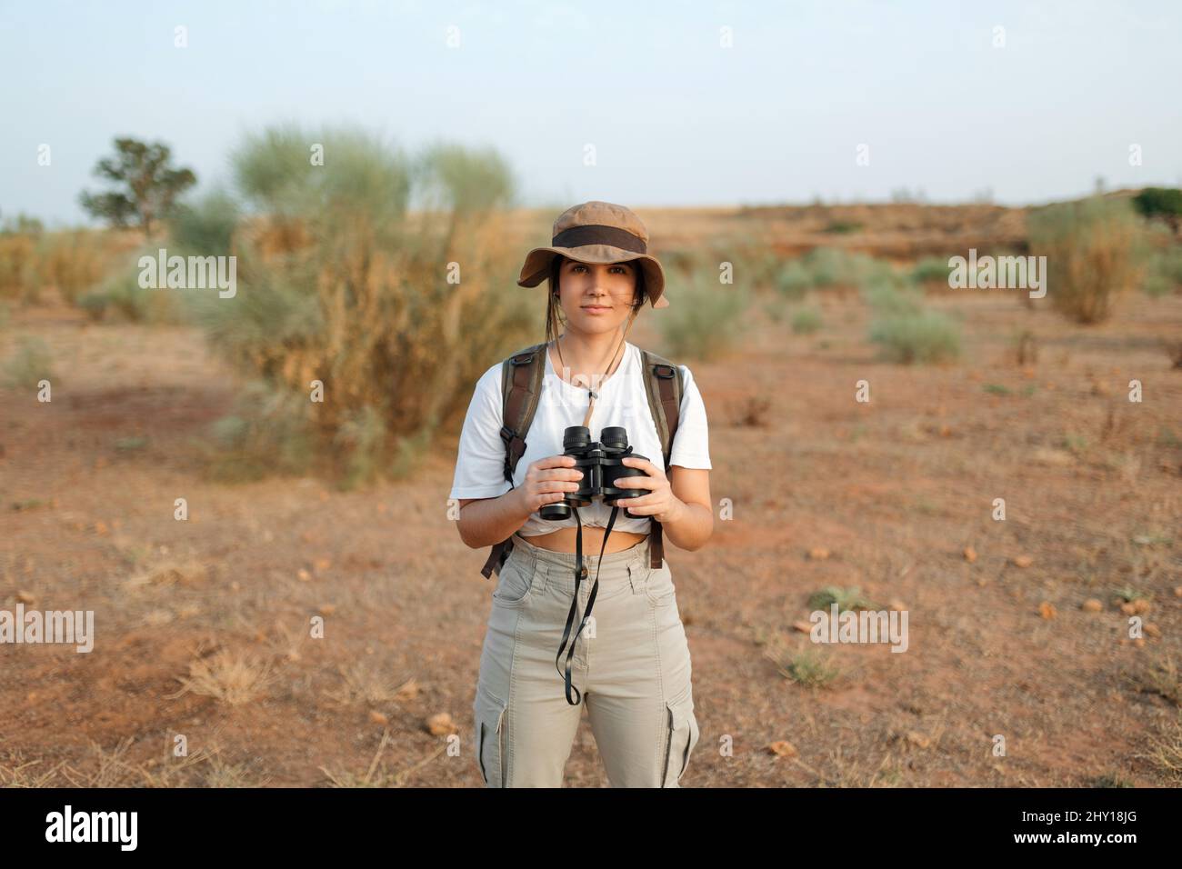 Female tourist in hat with binoculars standing looking at camera in desert area with green ...