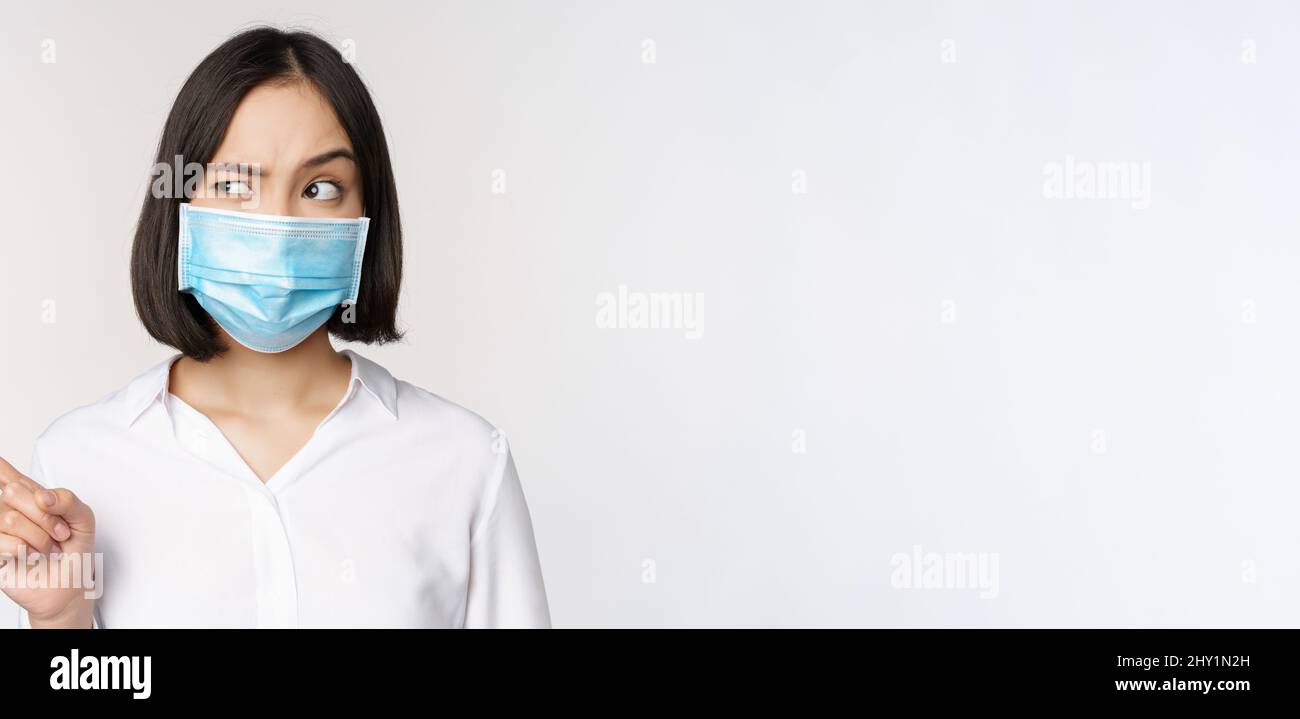 Image of young asian woman in medical face mask, pointing left and ...
