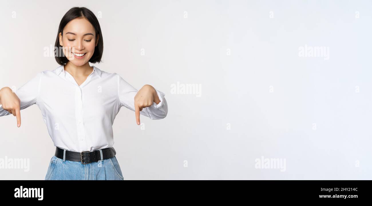 Portrait of happy asian woman pointing fingers down and looking below ...