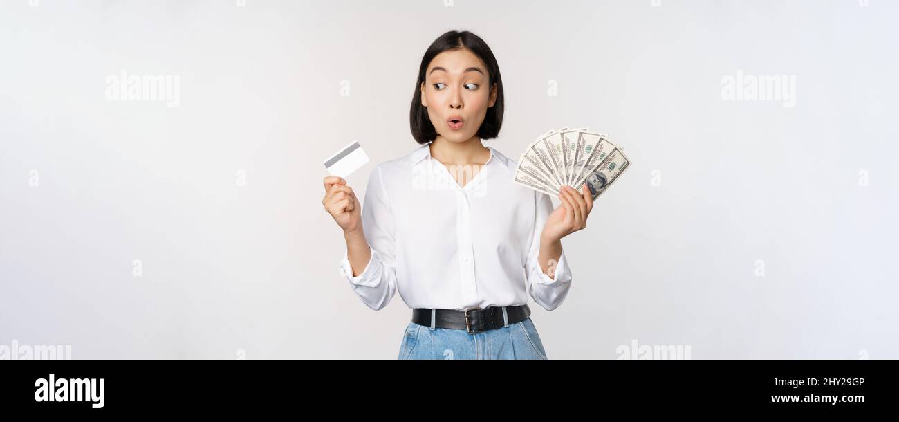 Excited korean girl looking at credit card, holding money cash, posing ...