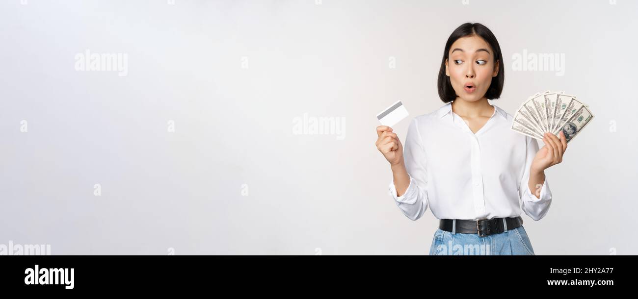 Excited korean girl looking at credit card, holding money cash, posing ...