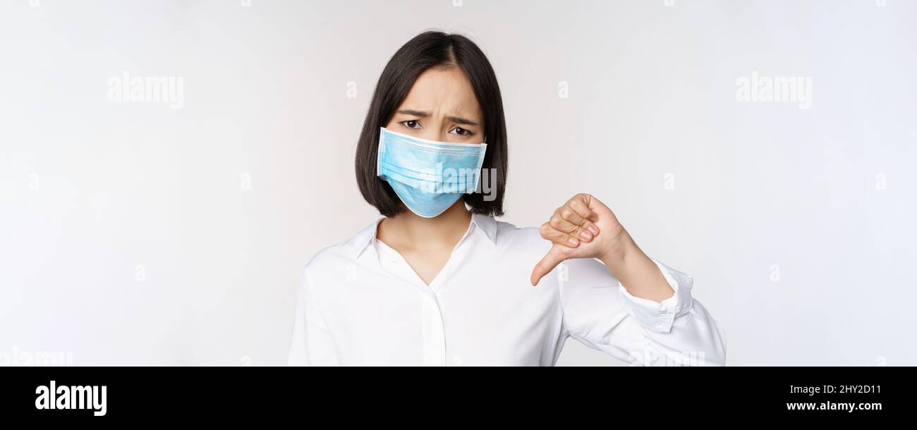 Portrait of asian woman in medical face mask showing thumbs down with ...