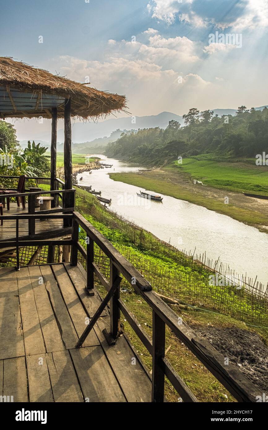 View of a hut with a wooden floor by a river, grass against a blue cloudy sky in Thanchi ...