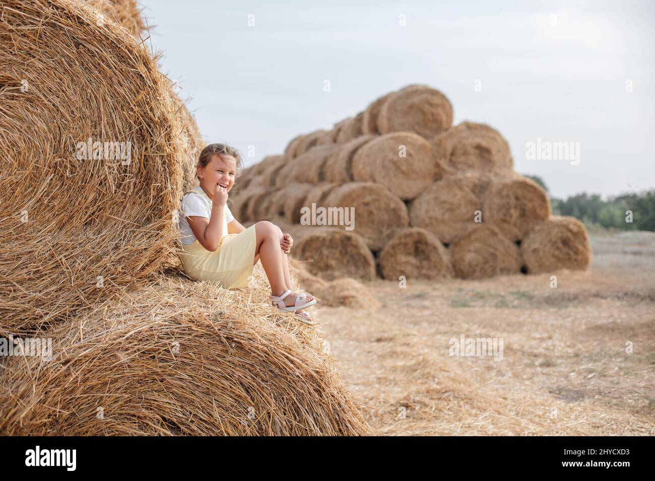 Excited little girl sitting on rolled haystack in sundress resting back ...