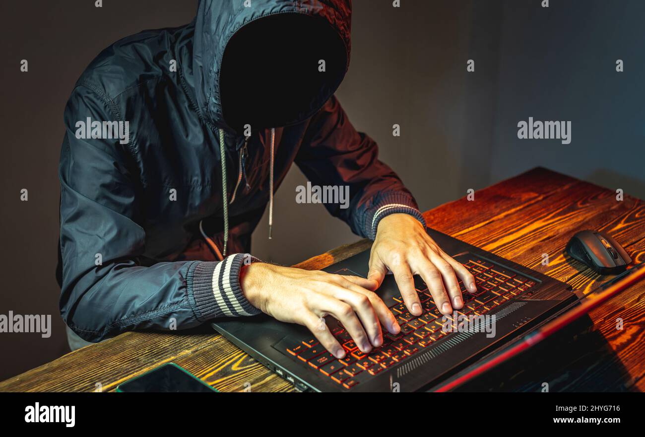 A hacker anonymous in a hood is typing on a laptop keyboard in a dark ...