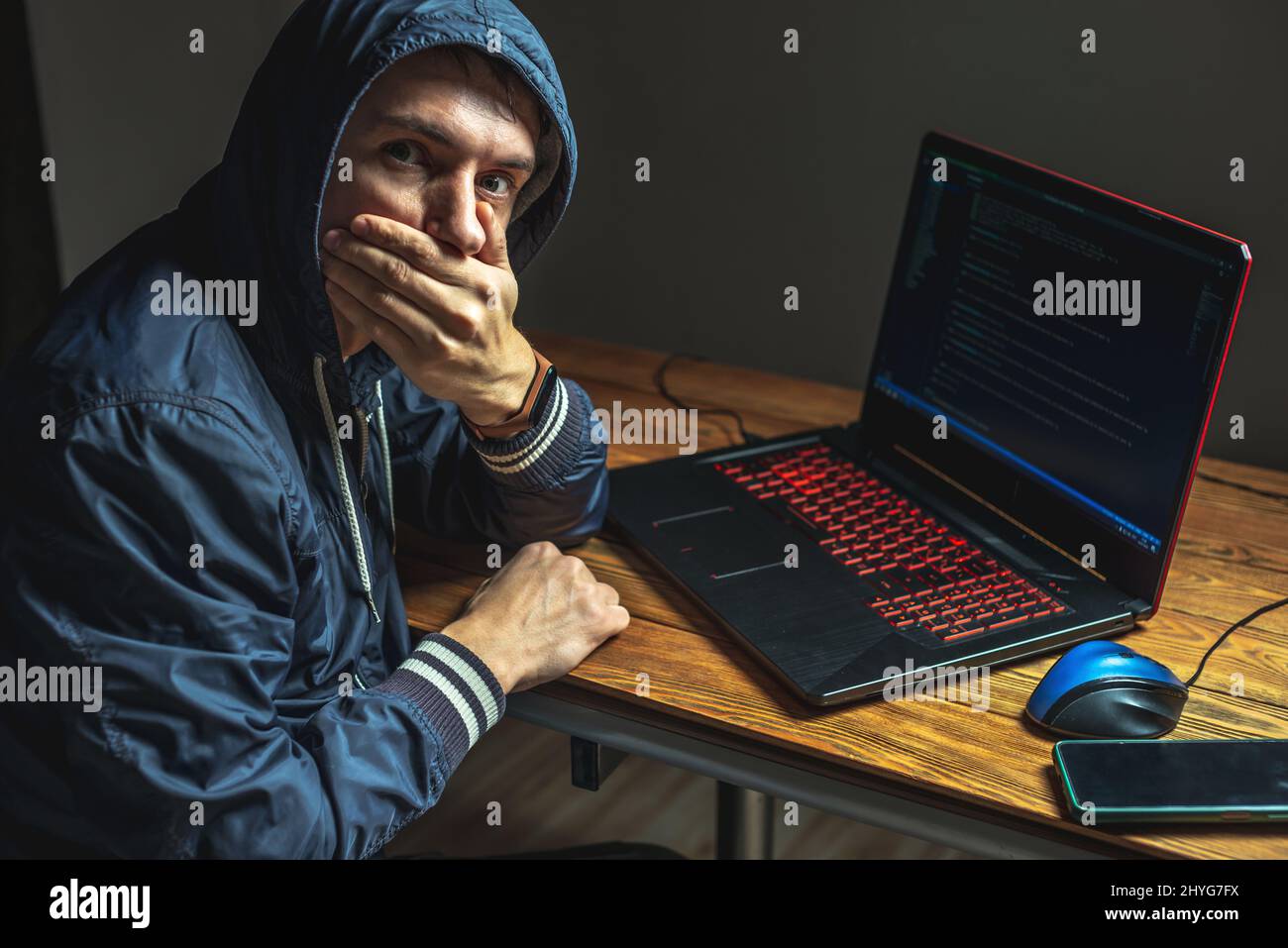 A hacker in a hood with a phone is typing on a laptop keyboard in a ...