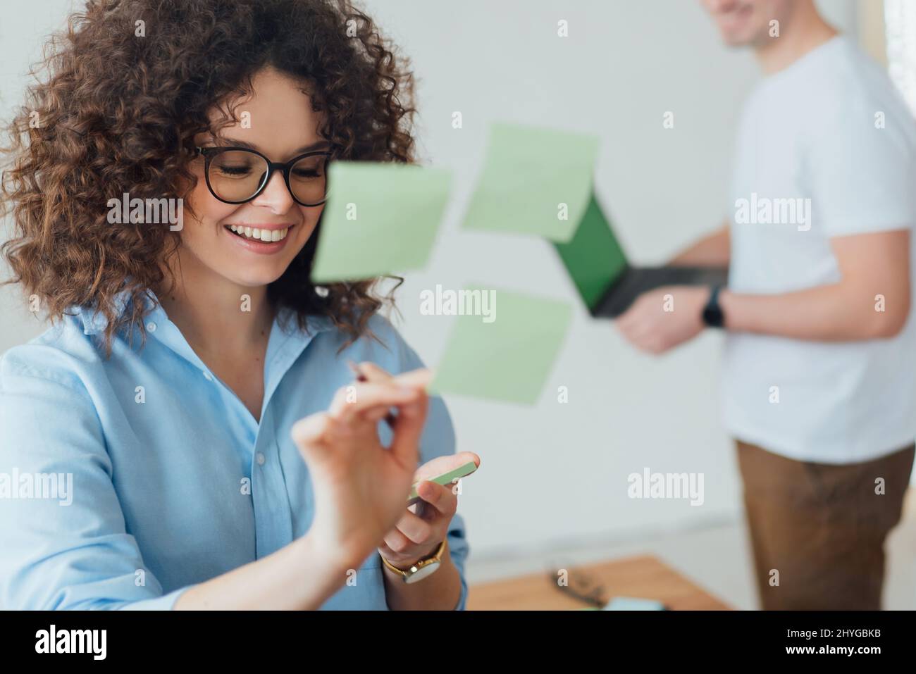 young curly woman with her colleague making notice by stickers. Team ...