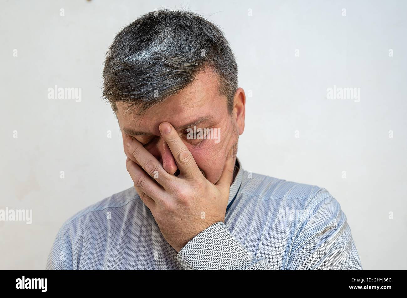 A mature man covers his face with his hand against the light wall. A ...