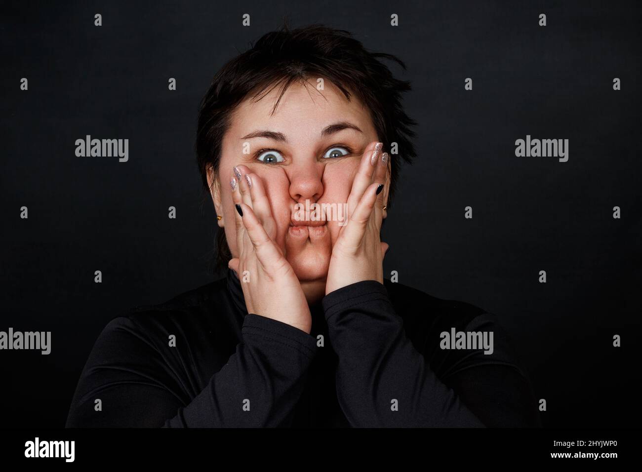 a brunette woman squeezed her face with her hands on a black background ...