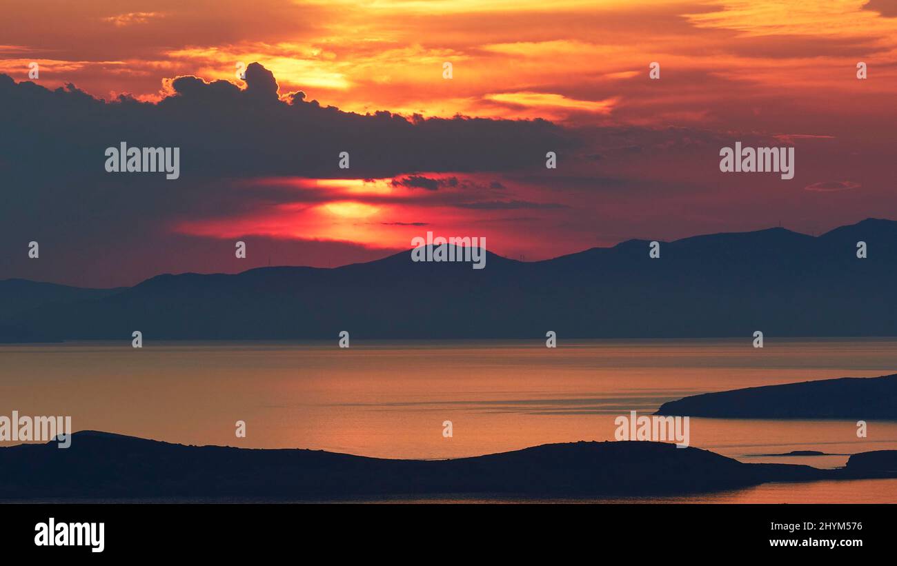 Sunset, sun big behind grey clouds and over a mountain range, headlands ...
